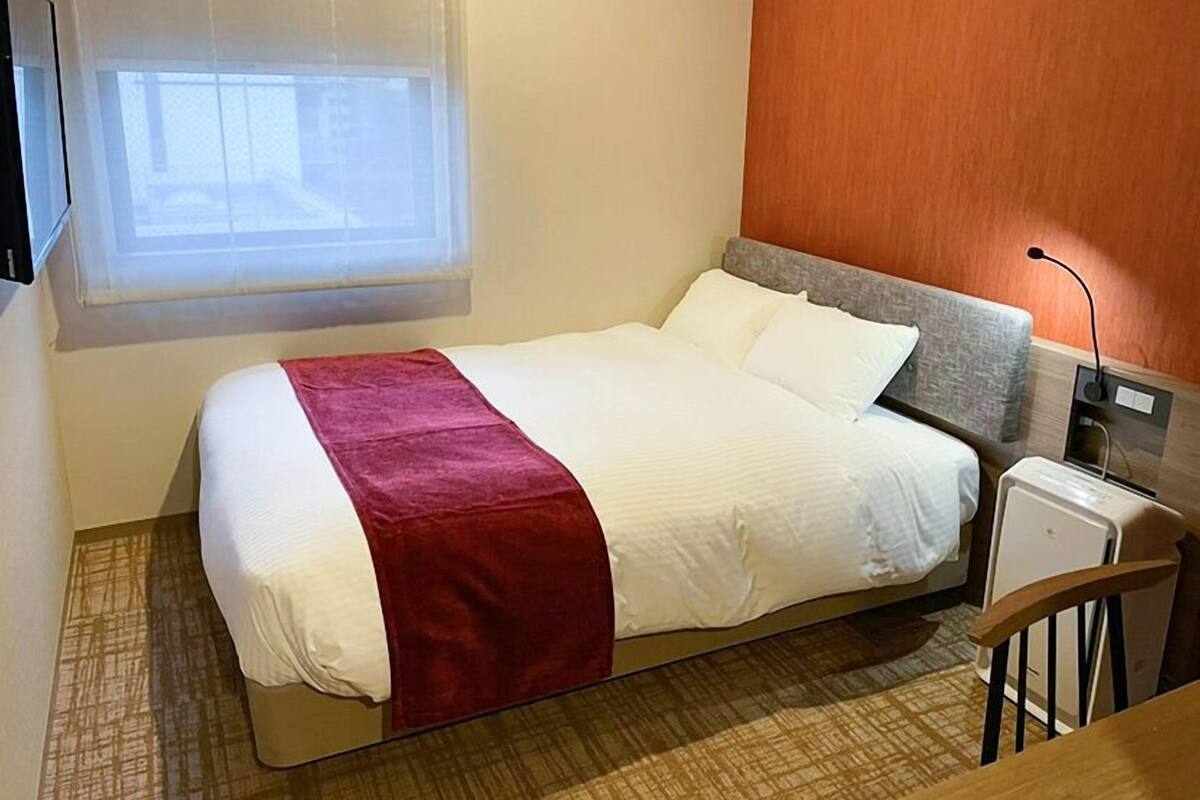 Quarto de hotel com cama, mesa de cabeceira, abajur e janela com persianas.