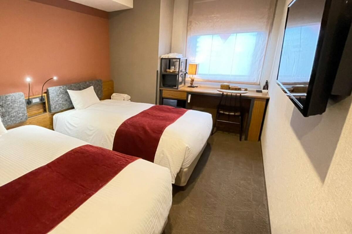 Quarto de hotel com duas camas, uma escrivaninha, uma televisão e uma janela.