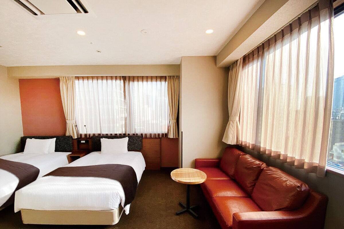 Quarto de hotel com duas camas, um sofá de couro vermelho, uma mesa redonda pequena e janelas grandes com cortinas leves.