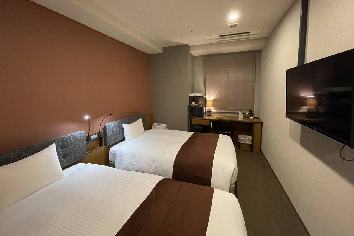Quarto de hotel com duas camas, uma escrivaninha com abajur, televisão e janela com cortinas.