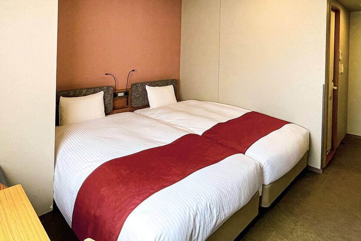 Quarto de hotel com uma cama grande, duas almofadas, cabeceira de madeira e um toque de vermelho na colcha.