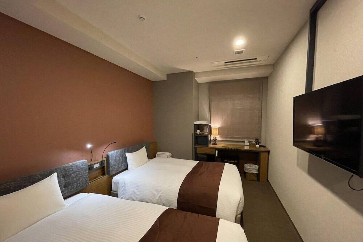 Quarto de hotel com duas camas, uma escrivaninha, uma televisão e uma janela.