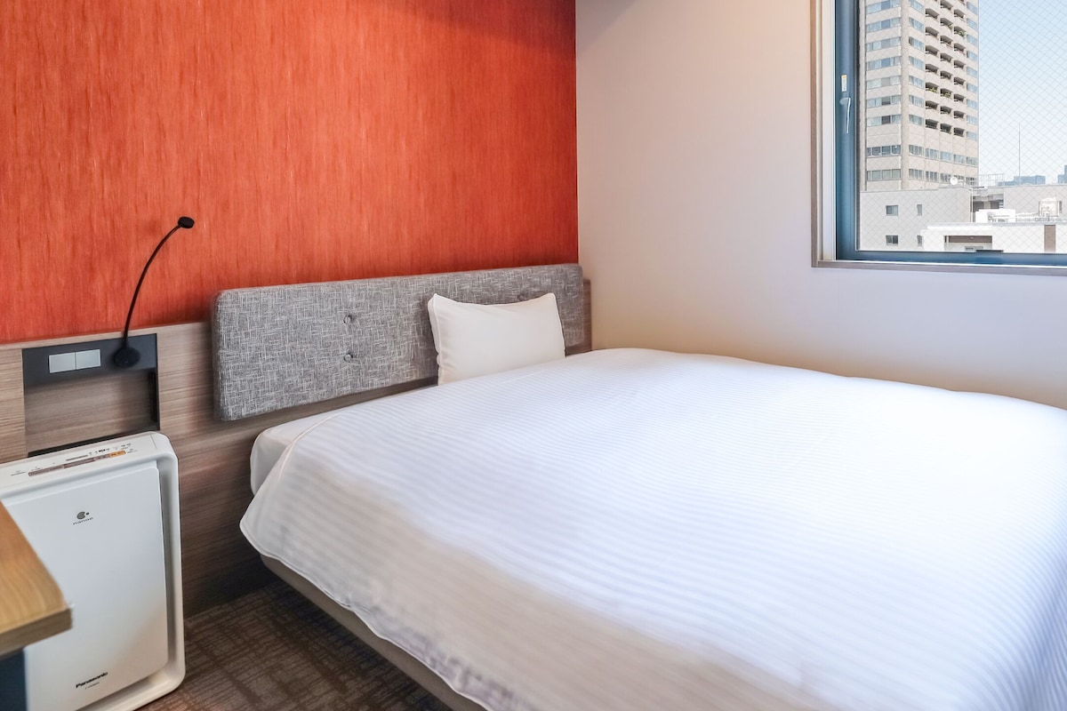 Quarto de hotel com uma cama, mesa de cabeceira com abajur, televisão na parede e janela com vista para edifícios.