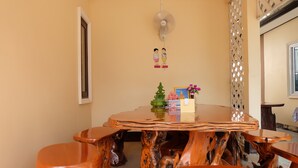 Uma área de refeições com uma mesa redonda de madeira e banquetas combinando, um ventilador fixado na parede e uma prateleira decorativa com pequenos objetos.