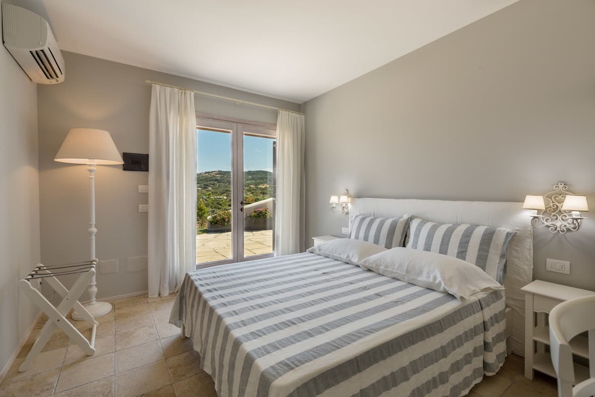 Quarto com cama, mesa de cabeceira, luminária, cadeira e vista para o exterior.