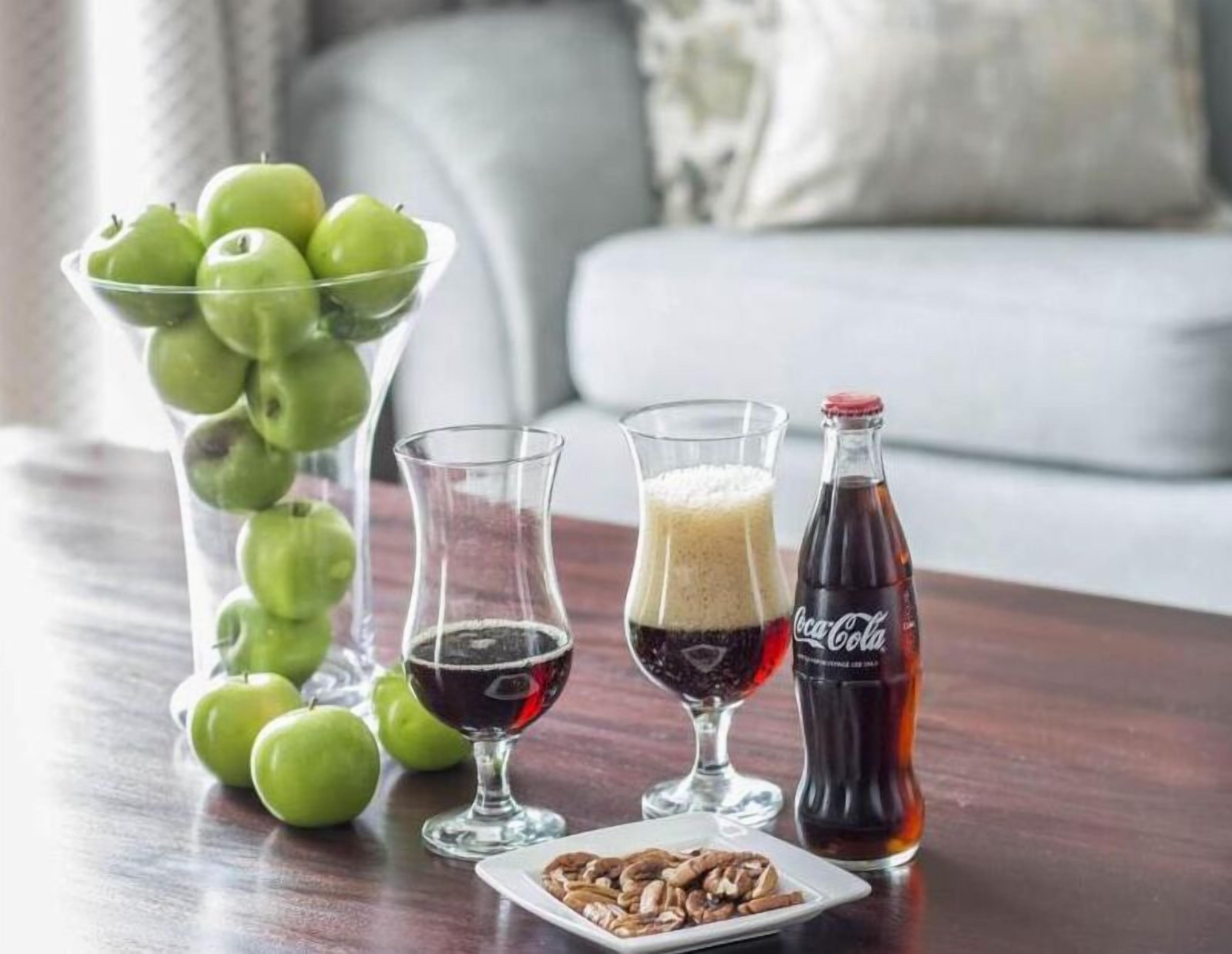 Uma mesa com um copo de líquido escuro, uma garrafa de Coca-Cola, uma tigela de maçãs verdes e um prato de nozes pecan.