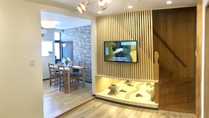 Uma sala de estar moderna com uma televisão de tela plana fixada em uma parede de painel de madeira, uma área de jantar com uma mesa e cadeiras de madeira, e uma escada com corrimão de madeira.
