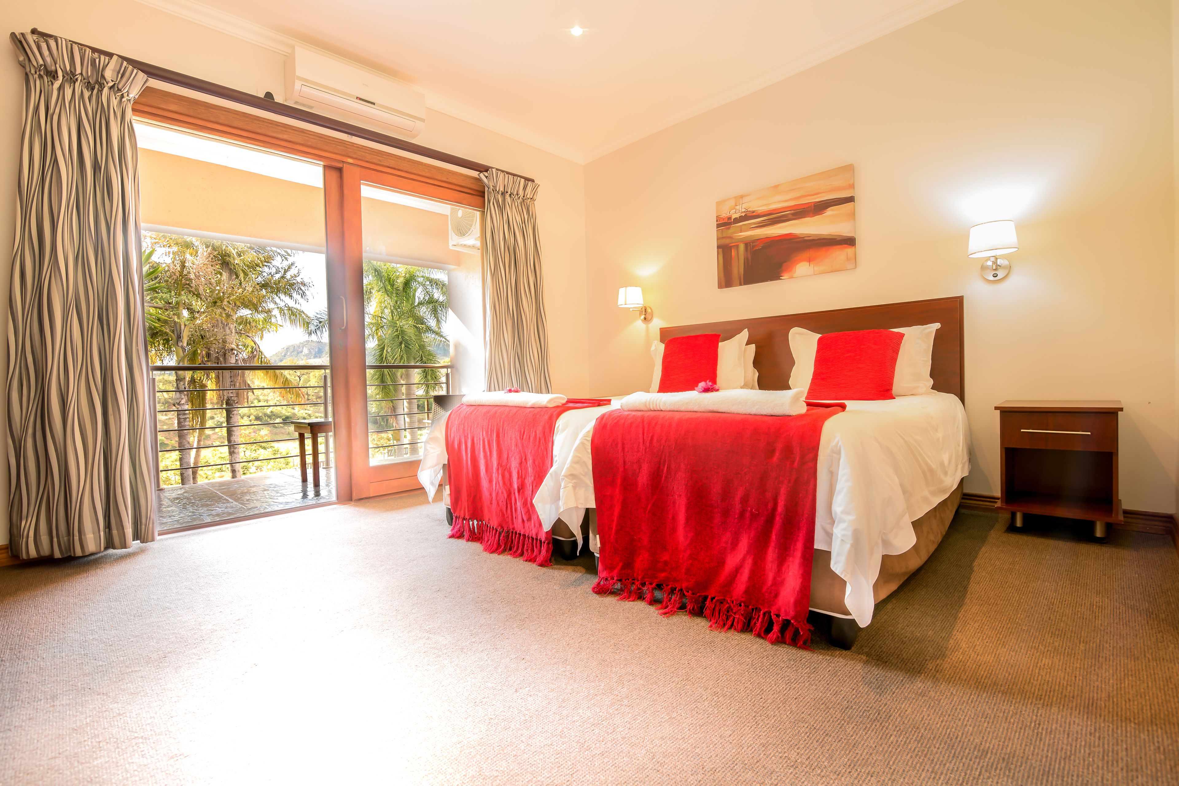 Quarto de hotel com uma cama grande, uma mesa de cabeceira, um quadro na parede e vista para palmeiras através das portas da varanda.