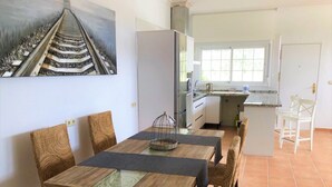 Uma área de refeições com mesa de madeira e cadeiras de vime, cozinha com armários brancos e um quadro de um trem na parede.
