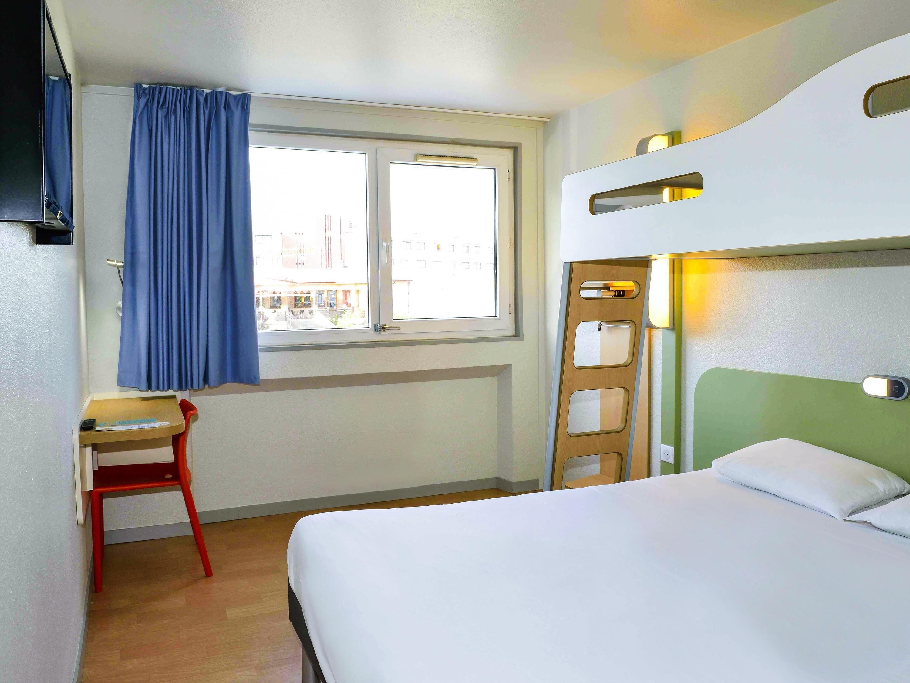 Quarto de hotel com cama, escrivaninha, cadeira, janela com cortinas e vista para prédios.