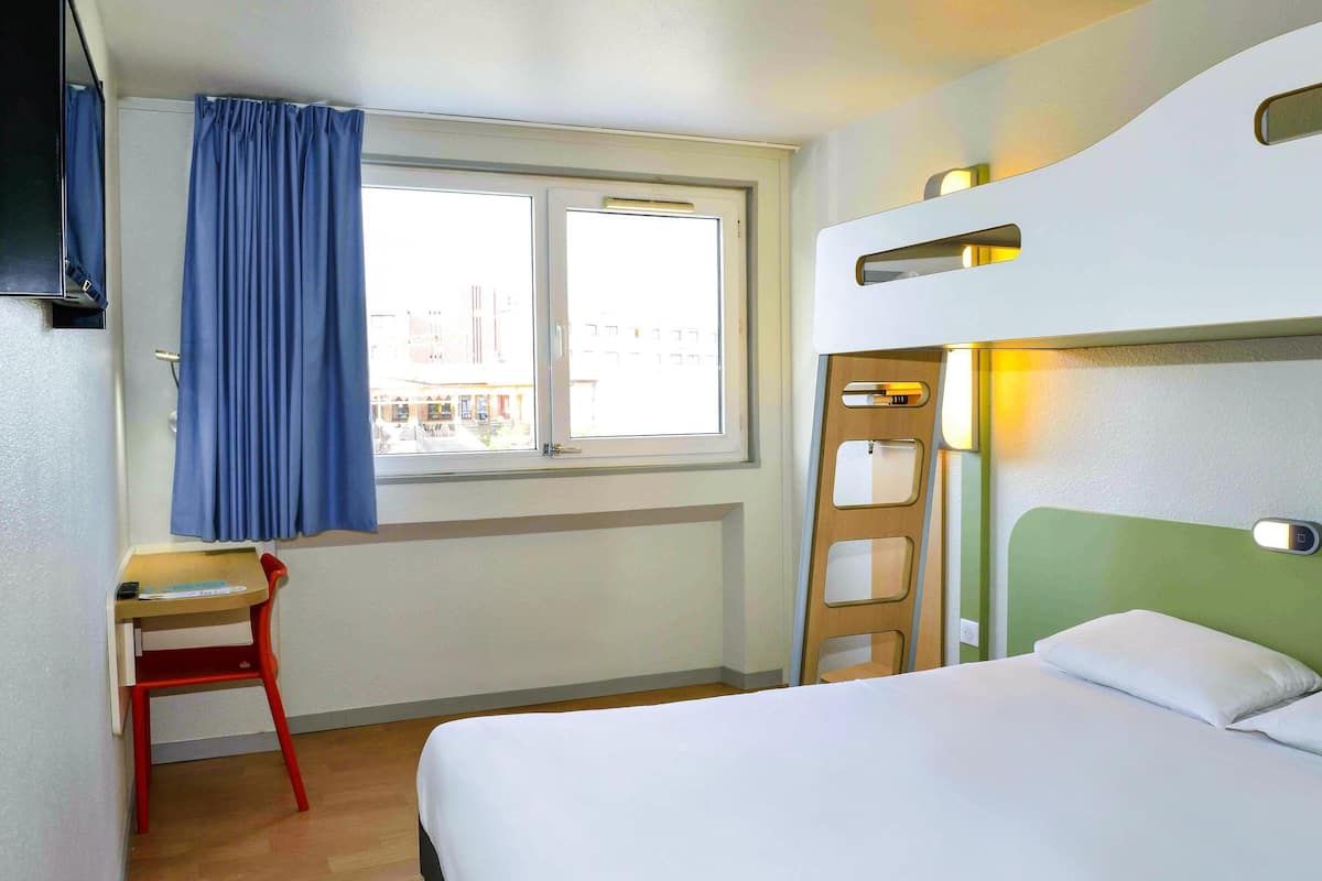 Quarto de hotel com cama, escrivaninha, cadeira, janela com cortinas e vista para prédios.