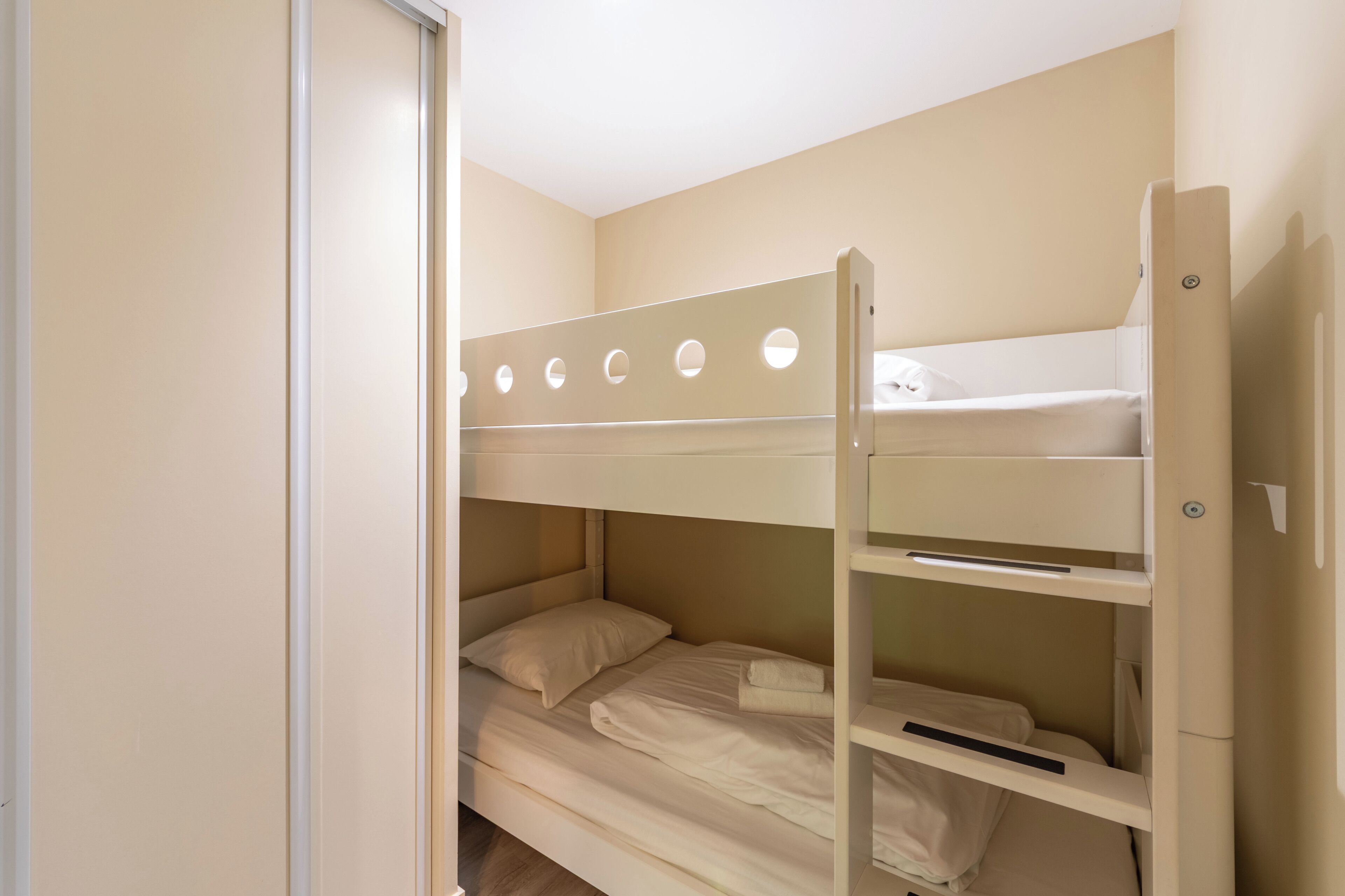 Uma cama box com peseira com recortes circulares, um espaço inferior com colchão e almofadas, e uma parede branca com uma porta.