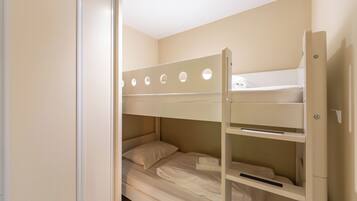 Uma cama box com peseira com recortes circulares, um espaço inferior com colchão e almofadas, e uma parede branca com uma porta.