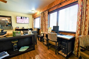 Quarto com piso de madeira, uma escrivaninha com computador, uma cadeira, televisão fixada na parede e uma janela com cortinas.