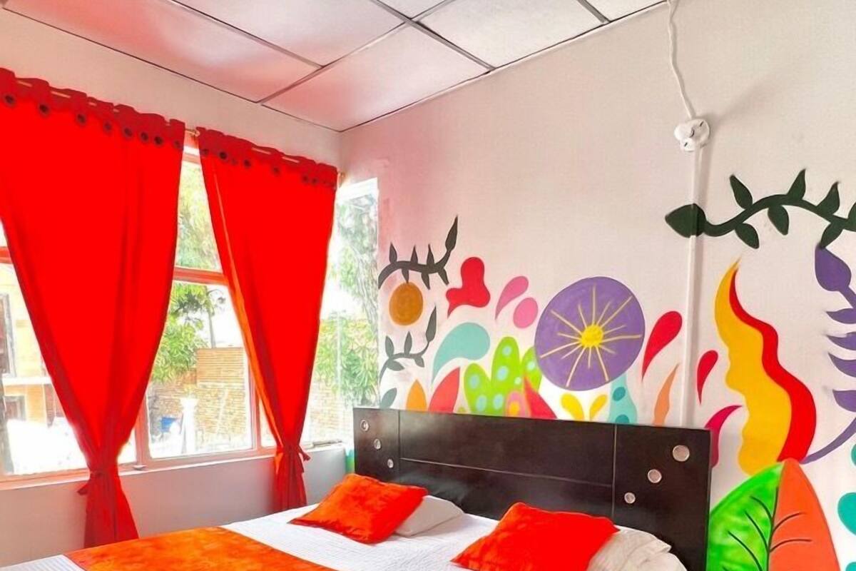 Quarto com cama, cortinas vermelhas, um mural colorido na parede e uma mesa de cabeceira.