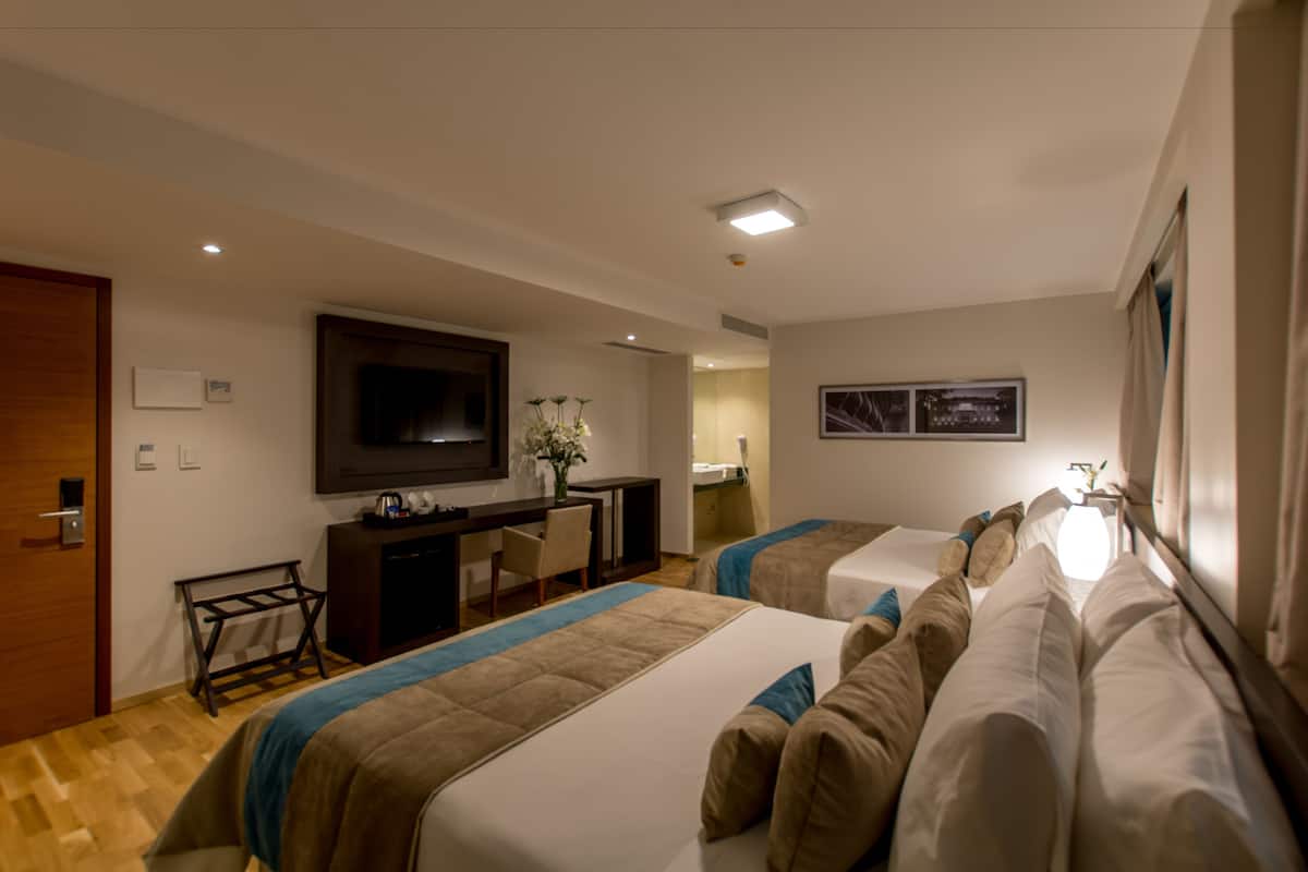 Quarto de hotel com duas camas, uma escrivaninha, televisão e janela com cortinas.