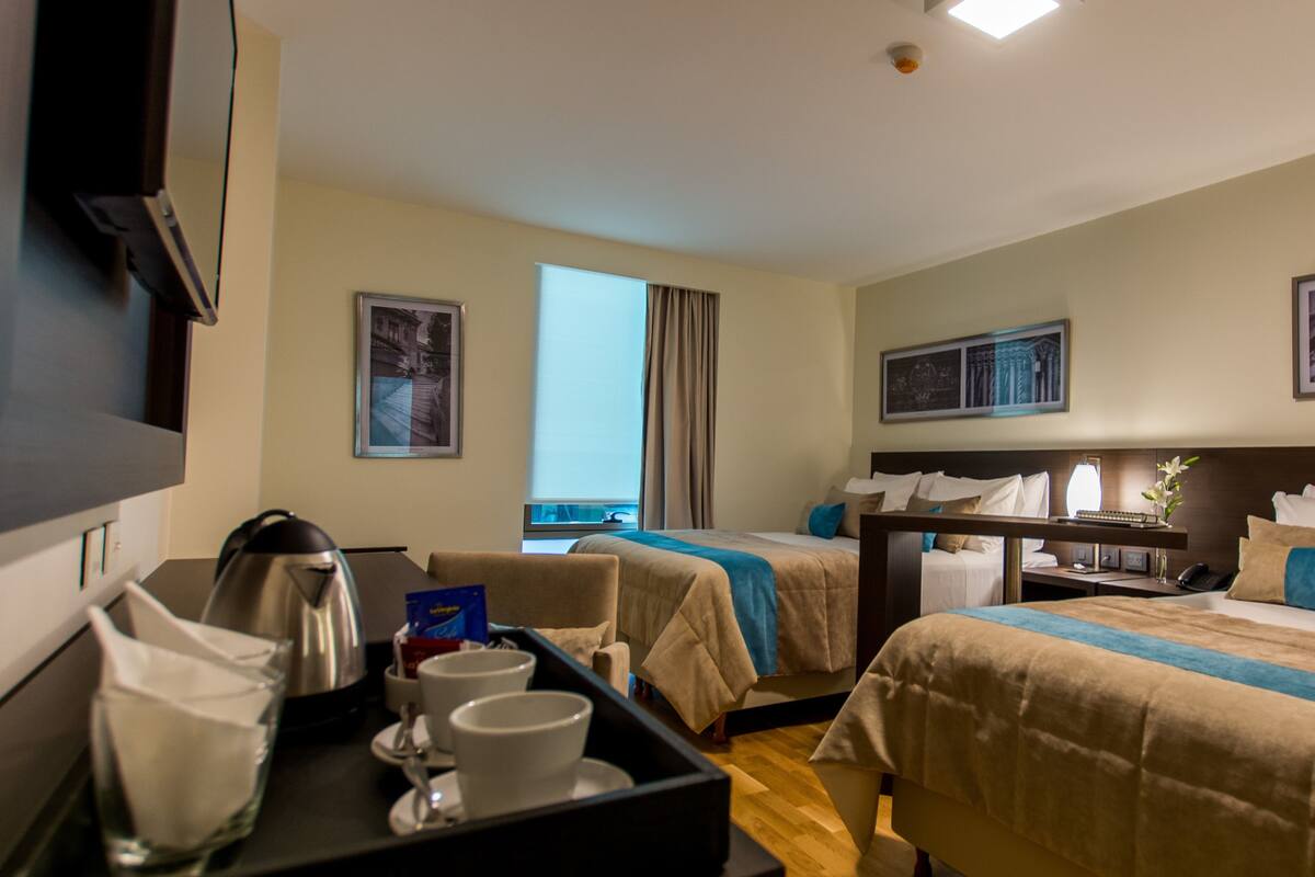 Quarto de hotel com duas camas, uma mesa de trabalho com chaleira e chávenas, televisão e janela com cortinas.