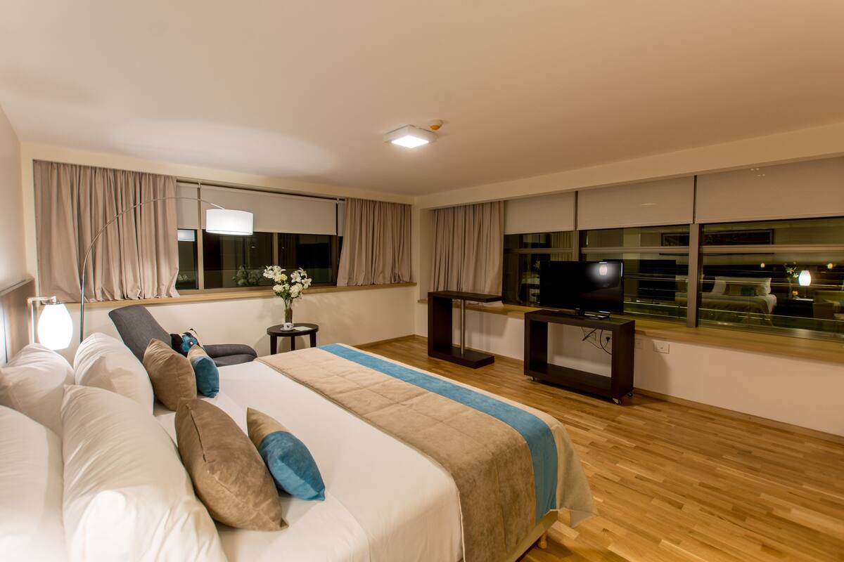 Quarto de hotel com uma cama grande, uma escrivaninha, uma televisão e uma janela com cortinas.