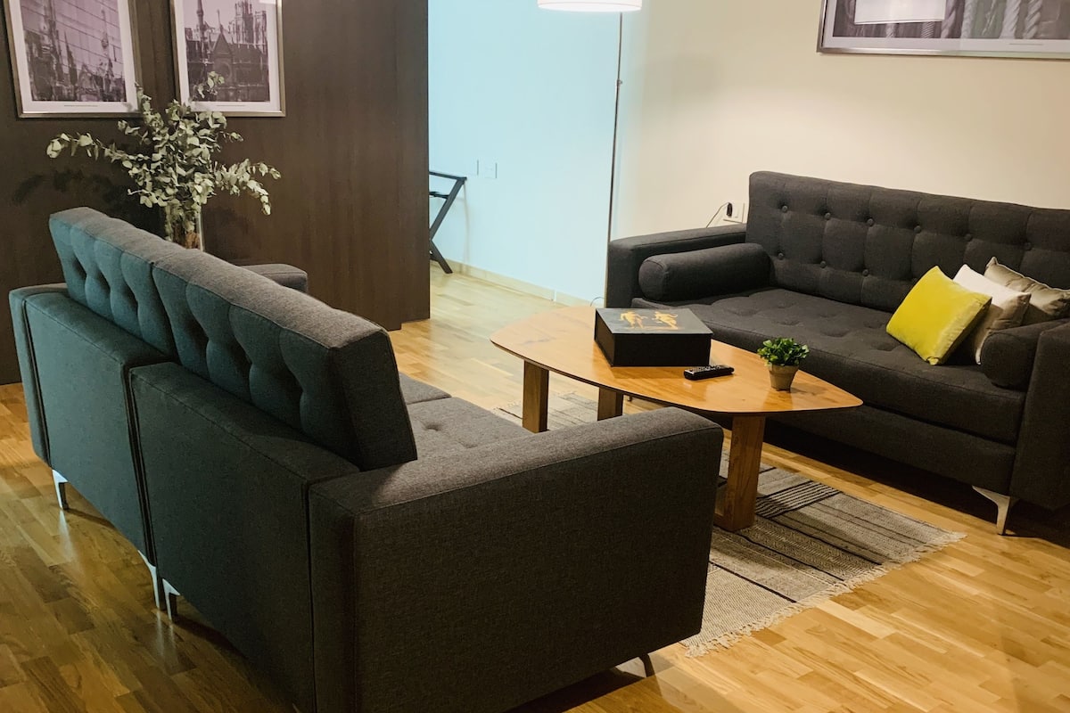 Uma sala de estar moderna com um sofá cinza escuro, uma poltrona coordenada e uma mesa de centro. Há quadros nas paredes, um abajur e uma janela grande ao fundo.