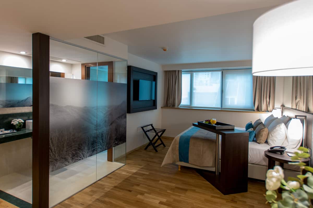 Quarto de hotel moderno com cama, escrivaninha, cadeira, janela ampla e espelho.
