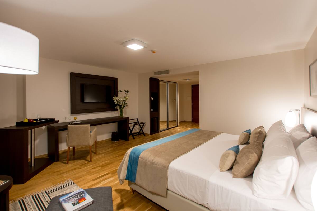 Quarto de hotel com uma cama, escrivaninha, cadeira, televisão e porta que dá para outro espaço.