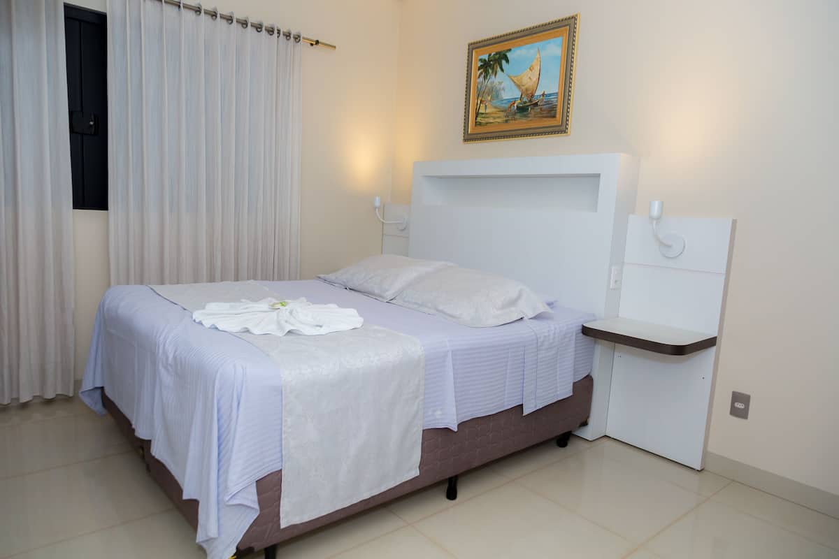 Cama bem arrumada com lençóis brancos, cabeceira e mesa de cabeceira com abajur.