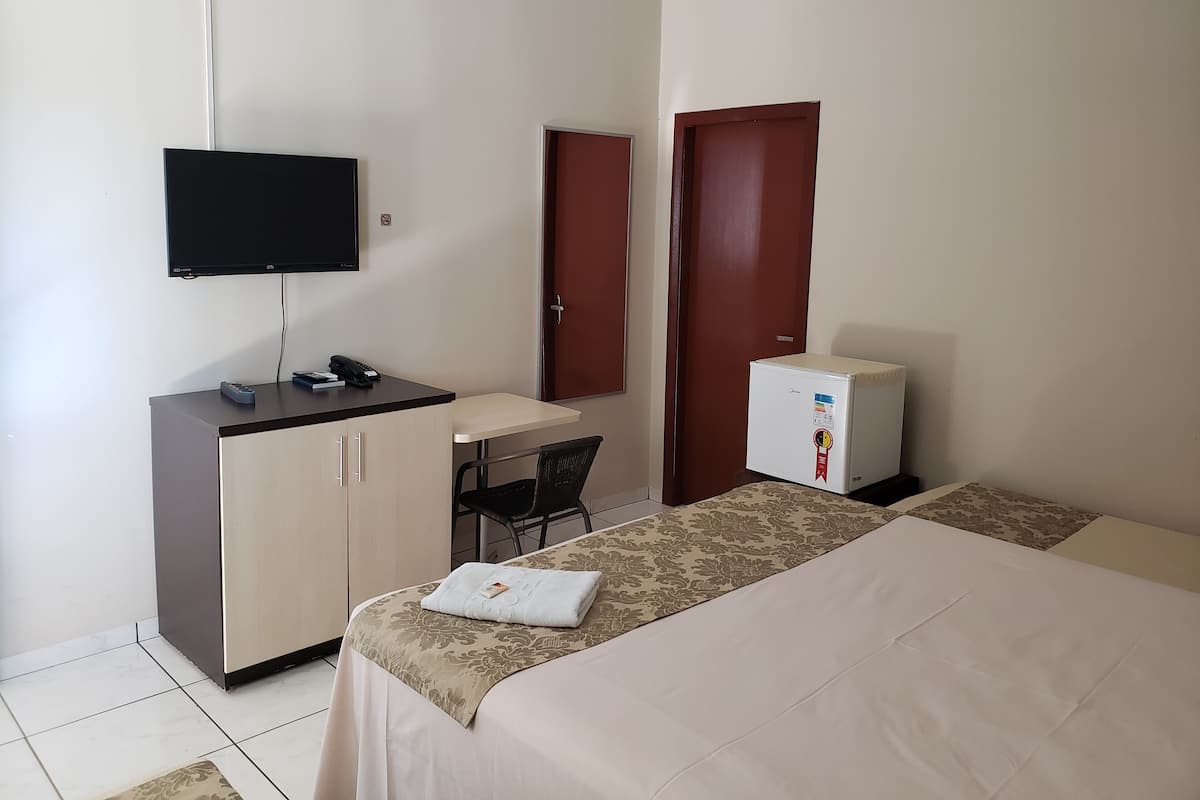 Quarto de hotel com cama, escrivaninha, cadeira, televisão e frigorífico.