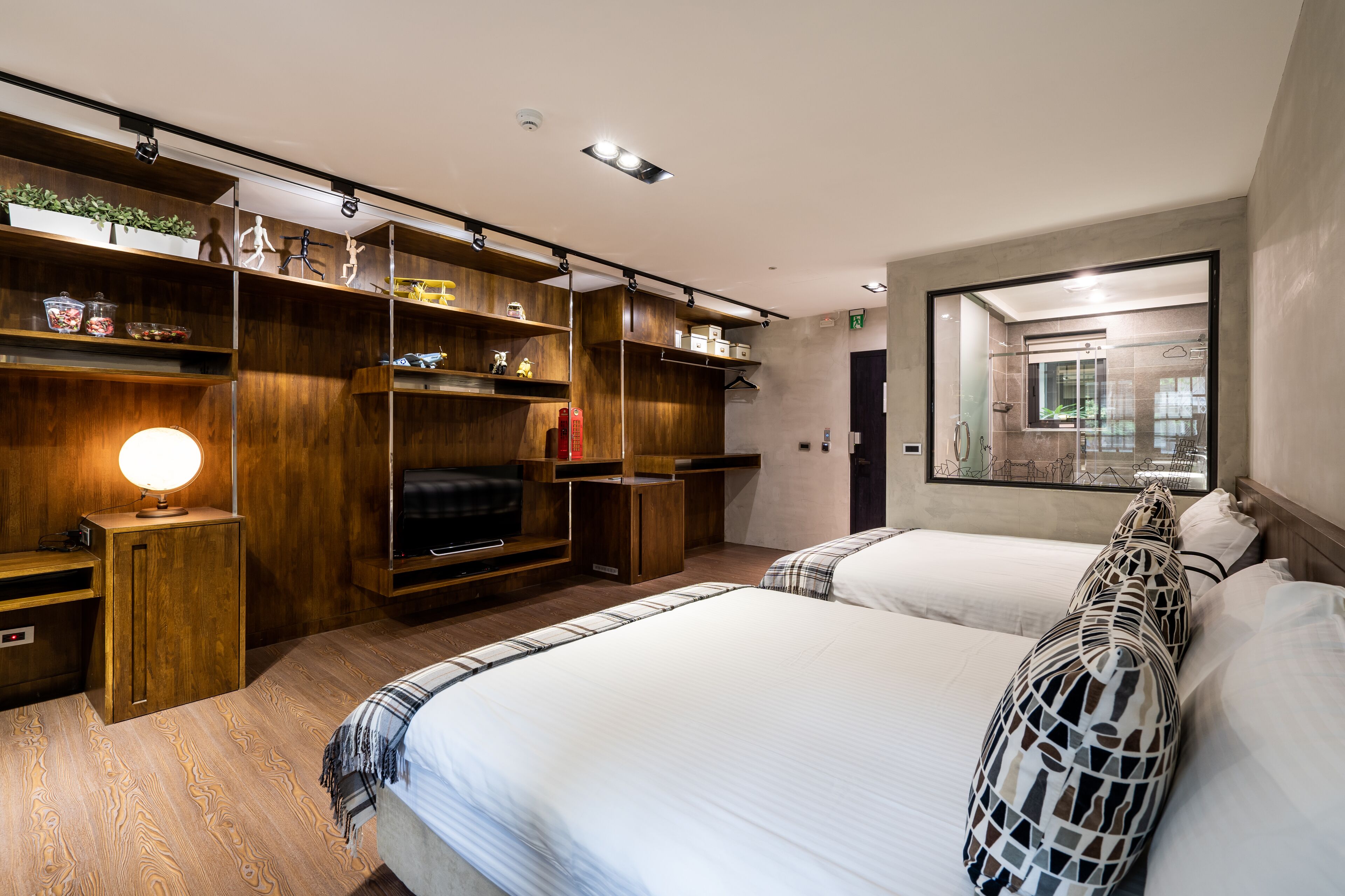 Quarto de hotel com duas camas, estantes de madeira com diversos objetos, uma televisão e uma janela grande com vista para o exterior.