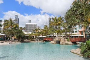 Área de piscina com uma ponte, palmeiras e edifícios ao fundo.