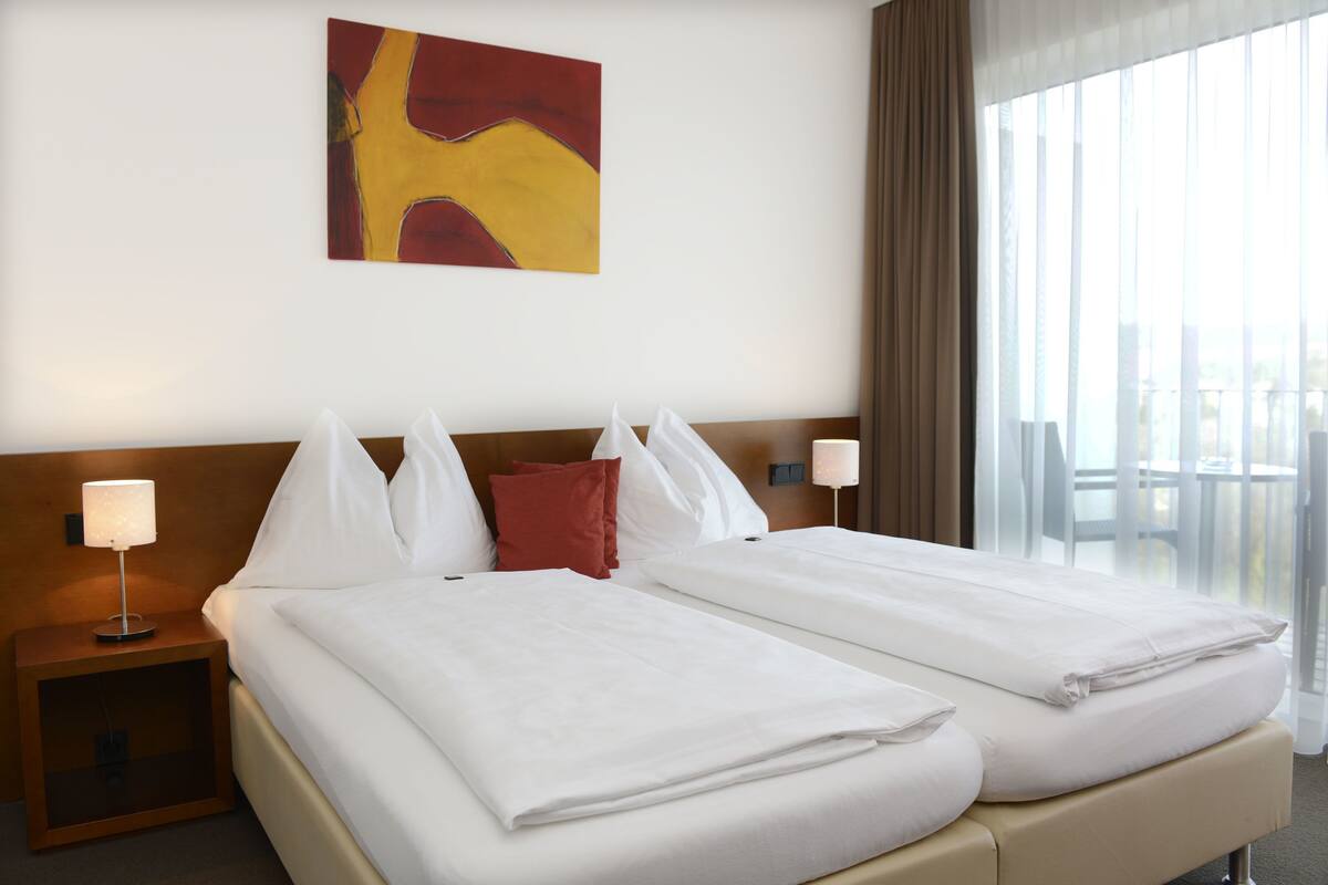 Quarto de hotel com duas camas, uma mesa de cabeceira com abajur, travesseiro vermelho e uma janela grande com vista.