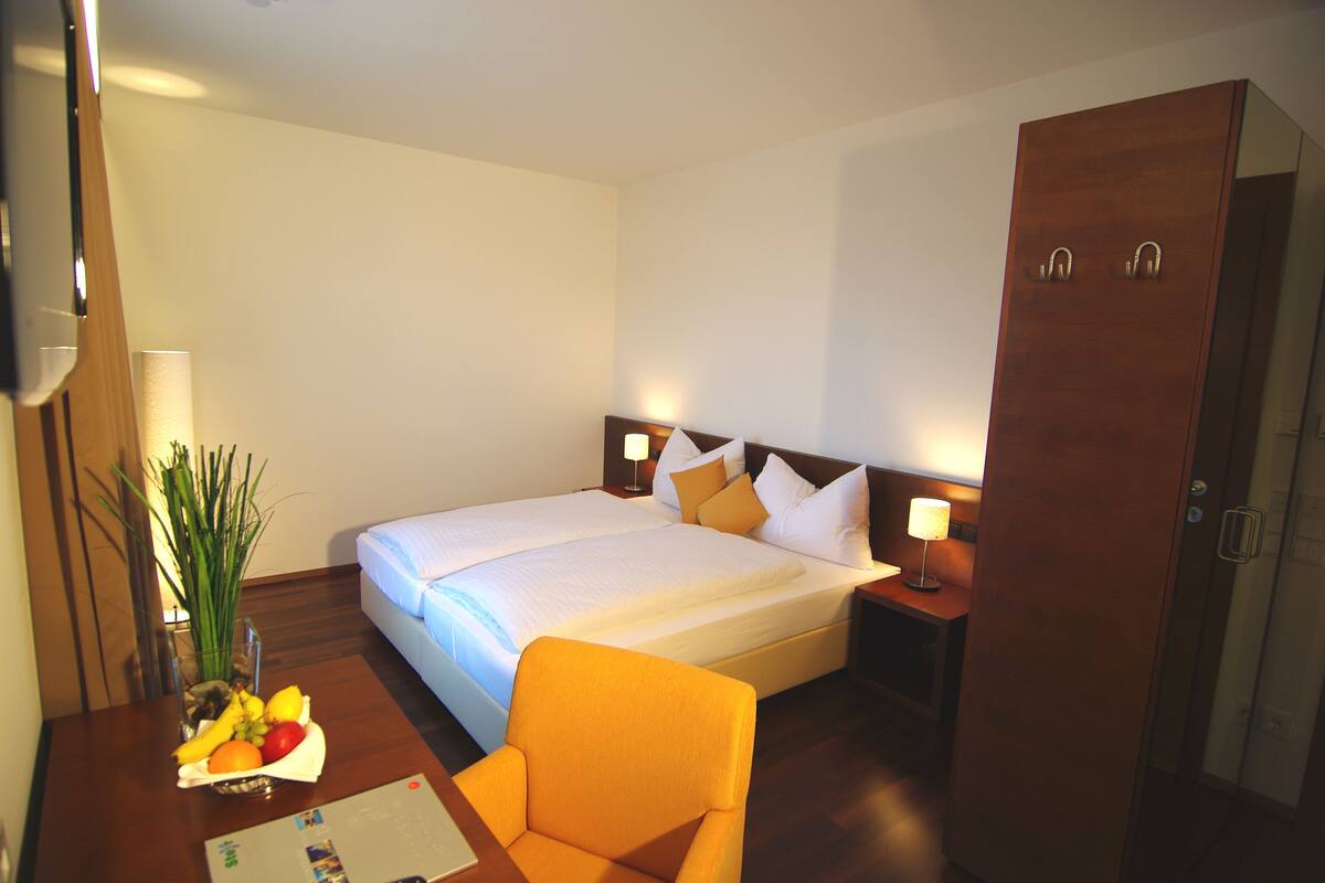 Quarto de hotel com uma cama grande, duas mesas de cabeceira, uma cadeira, uma escrivaninha com abajur e um guarda-roupa de madeira.