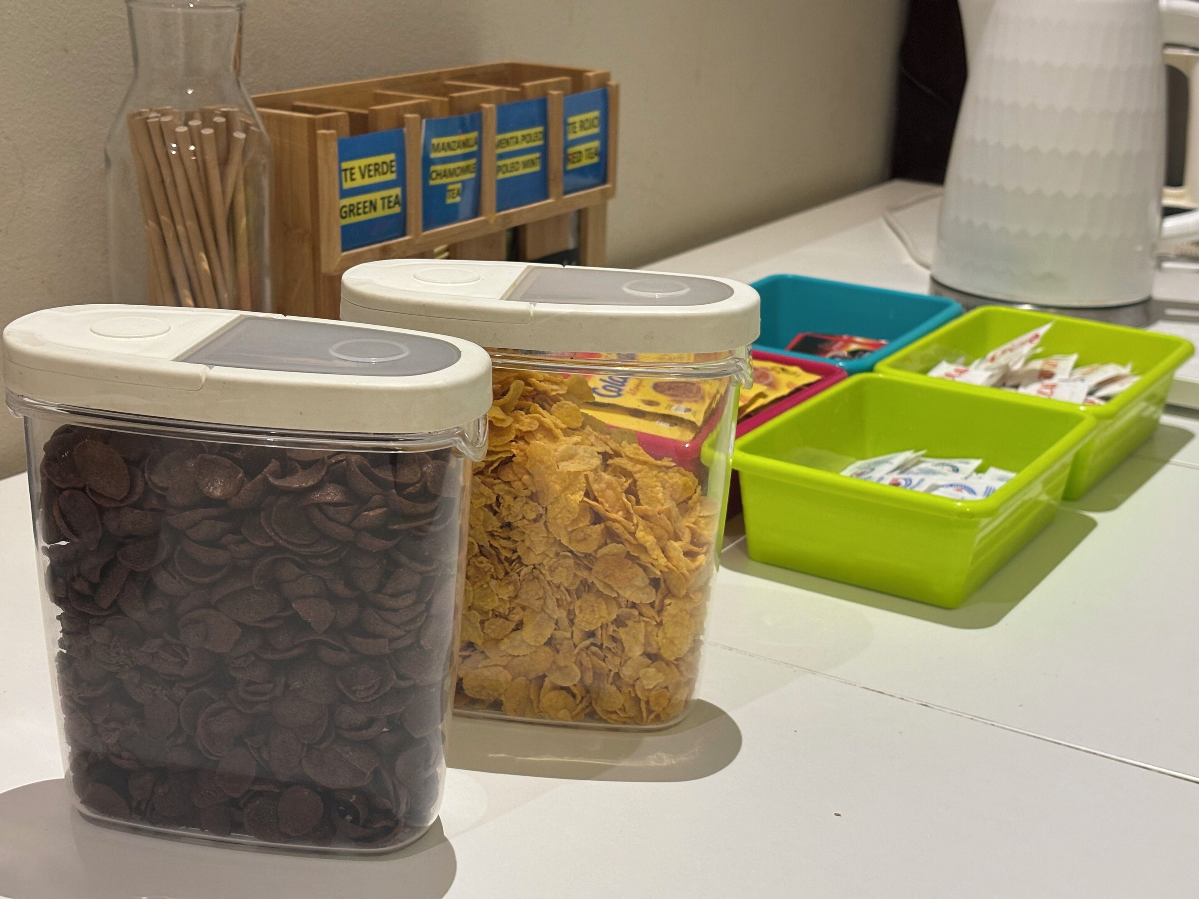 Dois recipientes de vidro com tampas, um com pedaços de chocolate e outro com flocos de cereais, e um conjunto de caixas de plástico coloridas com etiquetas.