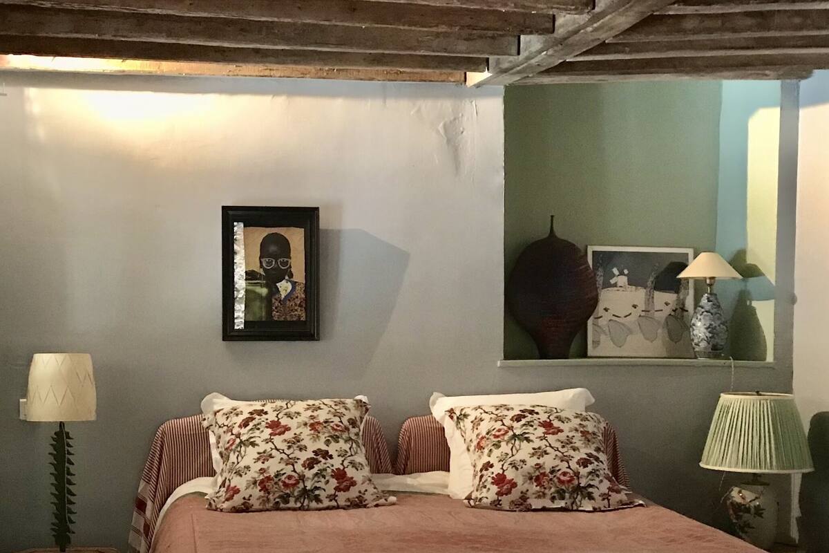Quarto com cama, almofadas, um quadro emoldurado, abajur e uma mesinha.