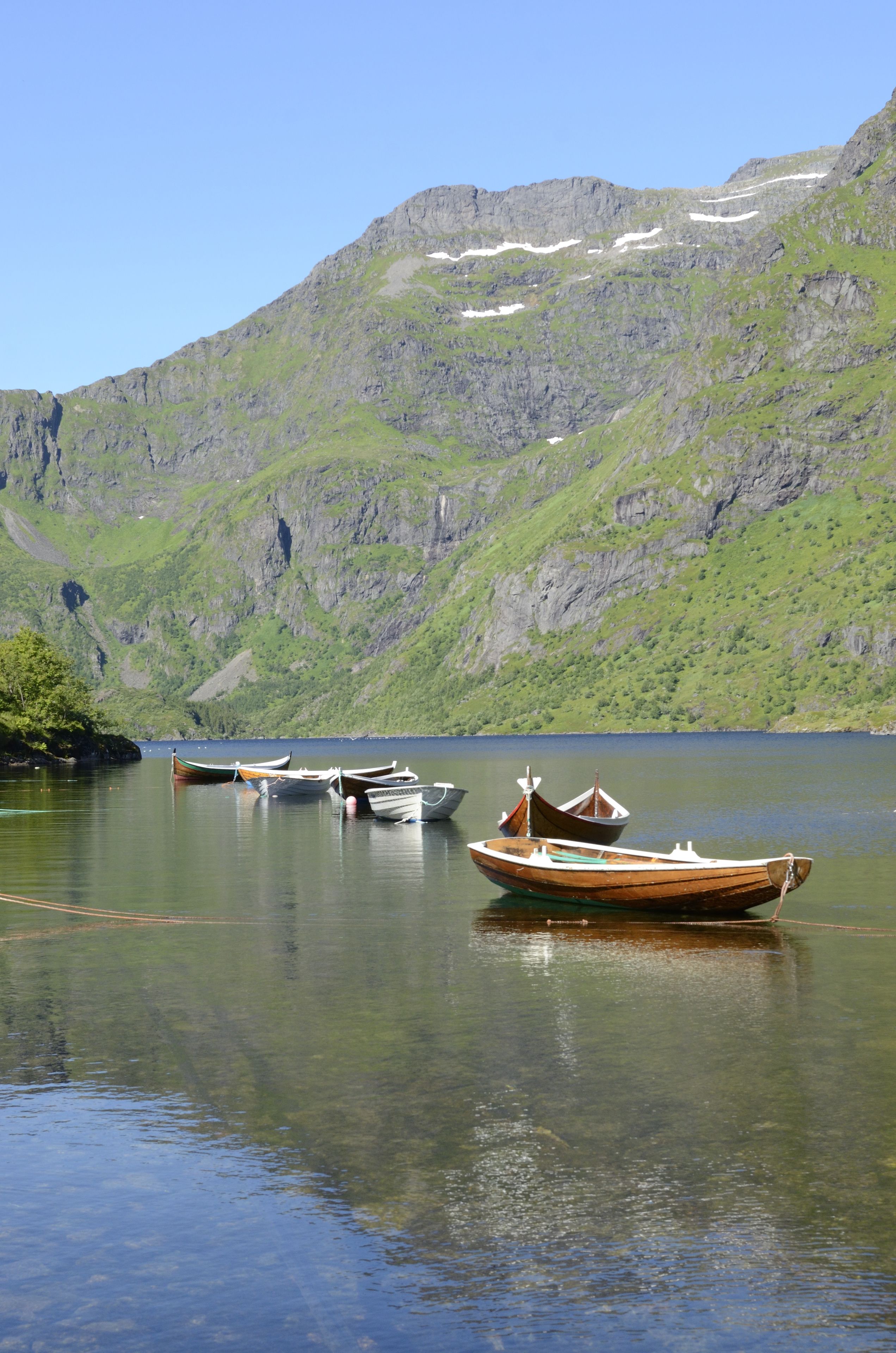 Um lago calmo com vários barcos atracados perto da margem, rodeado por colinas verdes e penhascos rochosos.