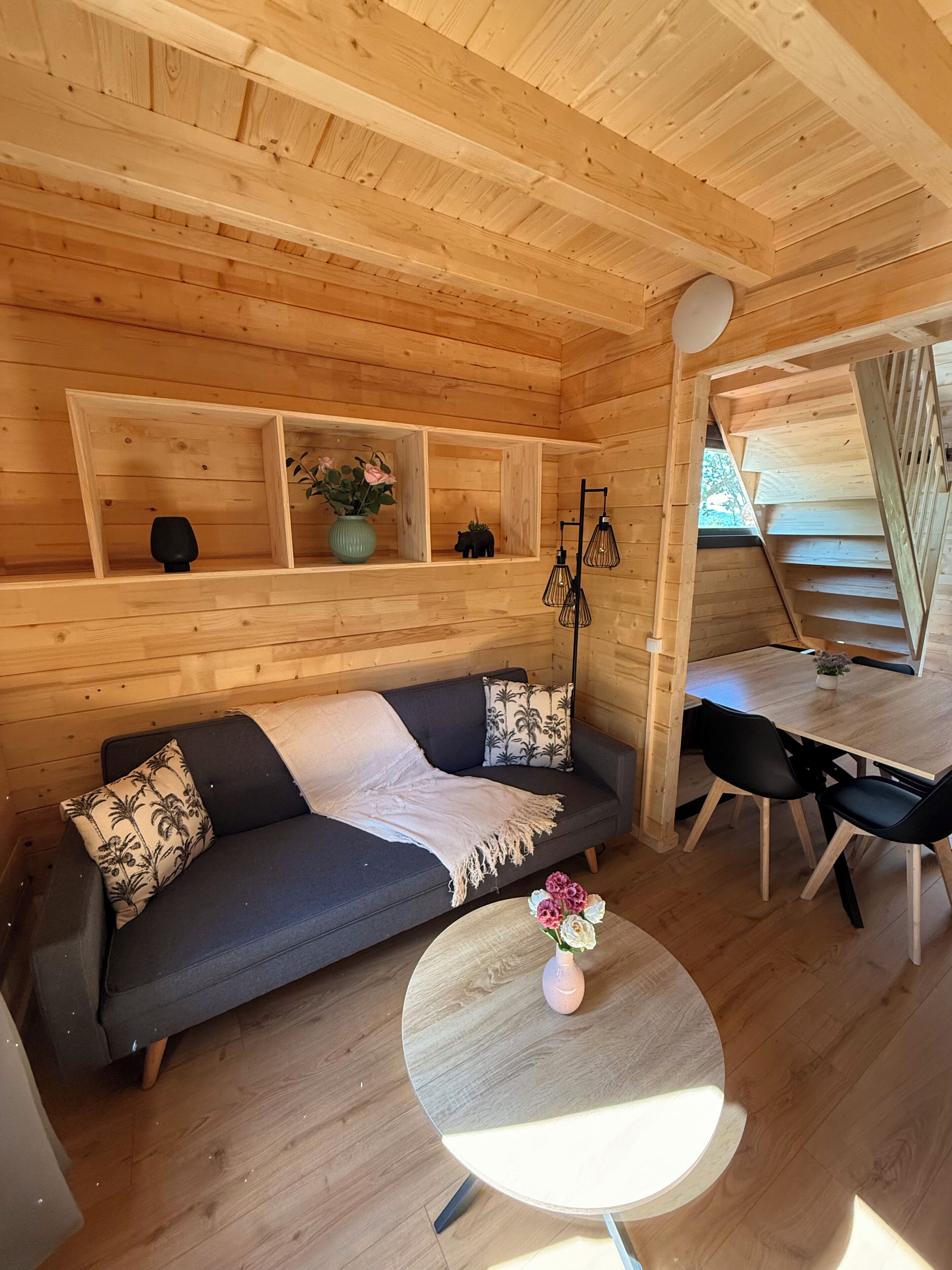 Interior acolhedor de uma cabana com um sofá cinza escuro, uma mesa redonda de madeira e uma área de jantar com cadeiras pretas.