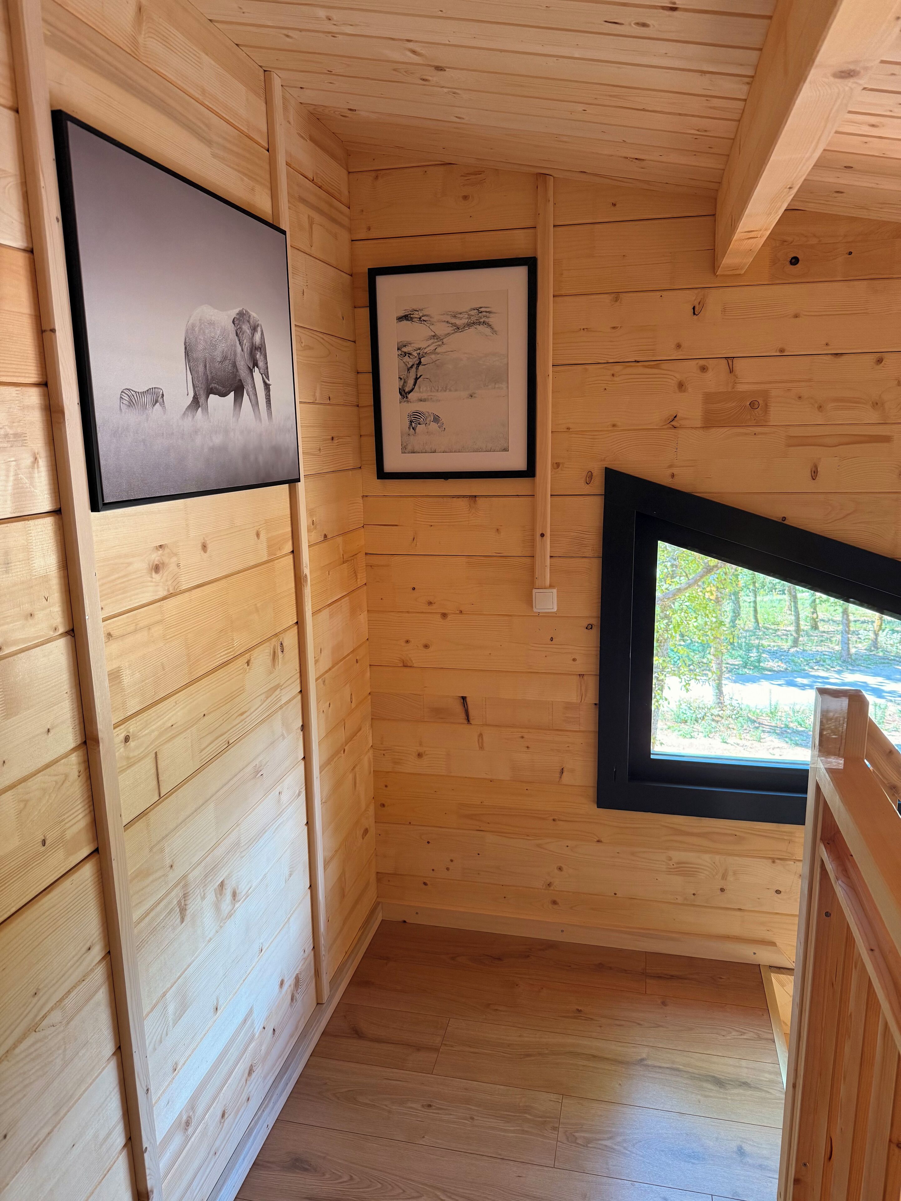 Quarto de cabana de madeira com duas imagens emolduradas nas paredes, uma janela com vista para vegetação e piso de madeira.