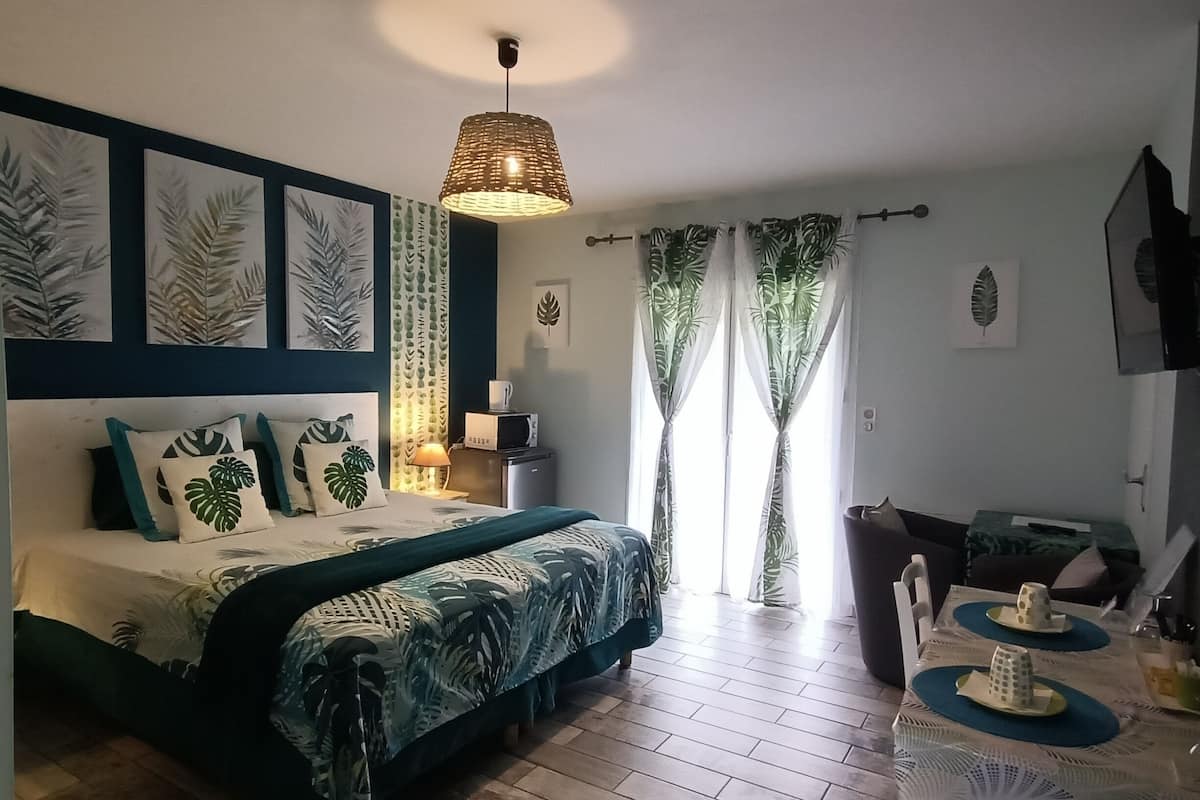 Quarto com cama, mesa de cabeceira, luminária e vista para o exterior através das cortinas.