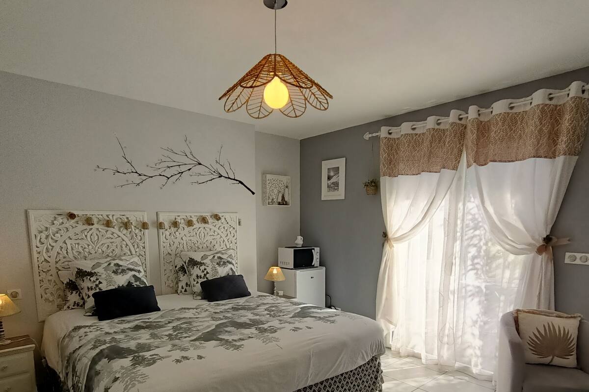 Um quarto com cama, mesa de cabeceira, cadeira, luminária, TV na parede e janela com cortinas.