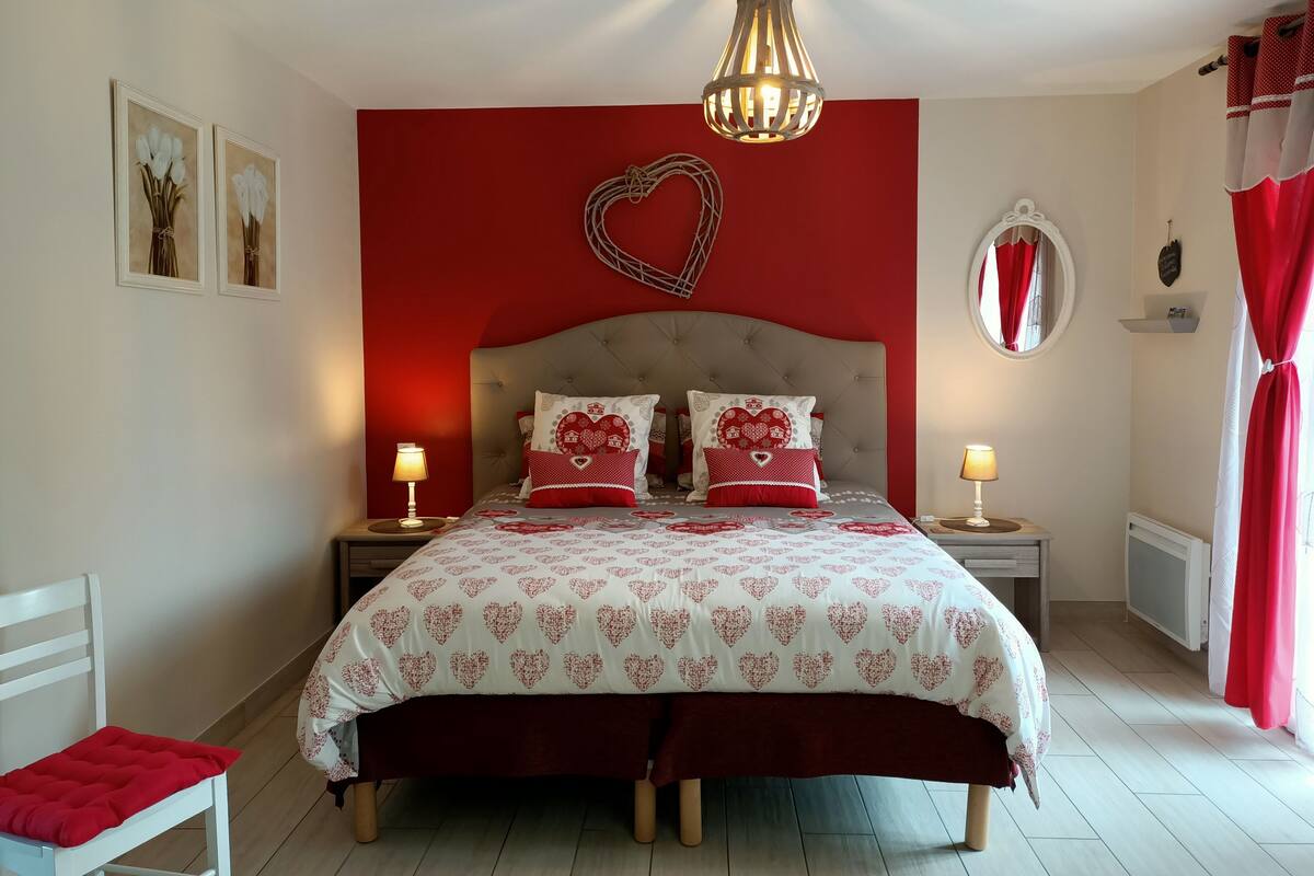 Quarto com parede de destaque vermelha, decoração em formato de coração, cabeceira estofada e cama com roupa de cama estampada em corações.