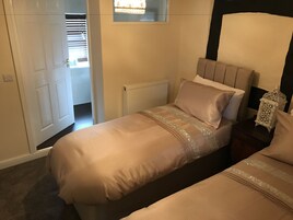 Quarto com duas camas, um radiador, uma janela com persianas e uma mesa de cabeceira com um candeeiro decorativo.