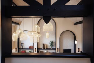 Uma área de recepção com luminárias trançadas penduradas, uma planta e uma estante com livros e objetos decorativos.