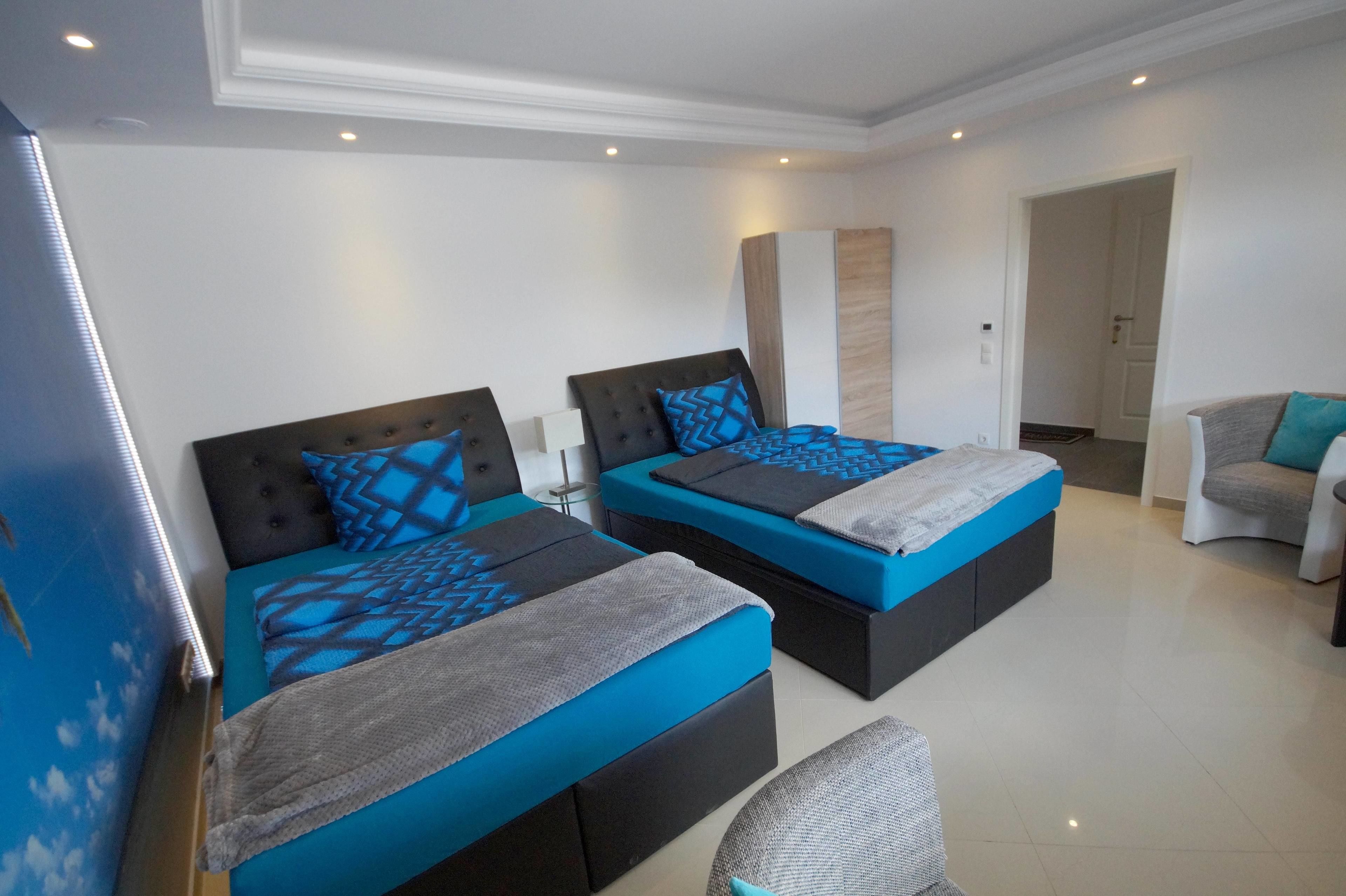 Quarto de hotel com duas camas, cada uma com colcha azul e cinzenta, um cobertor cinza e cabeceira preta. Há uma poltrona cinza e uma mesinha de centro com abajur.