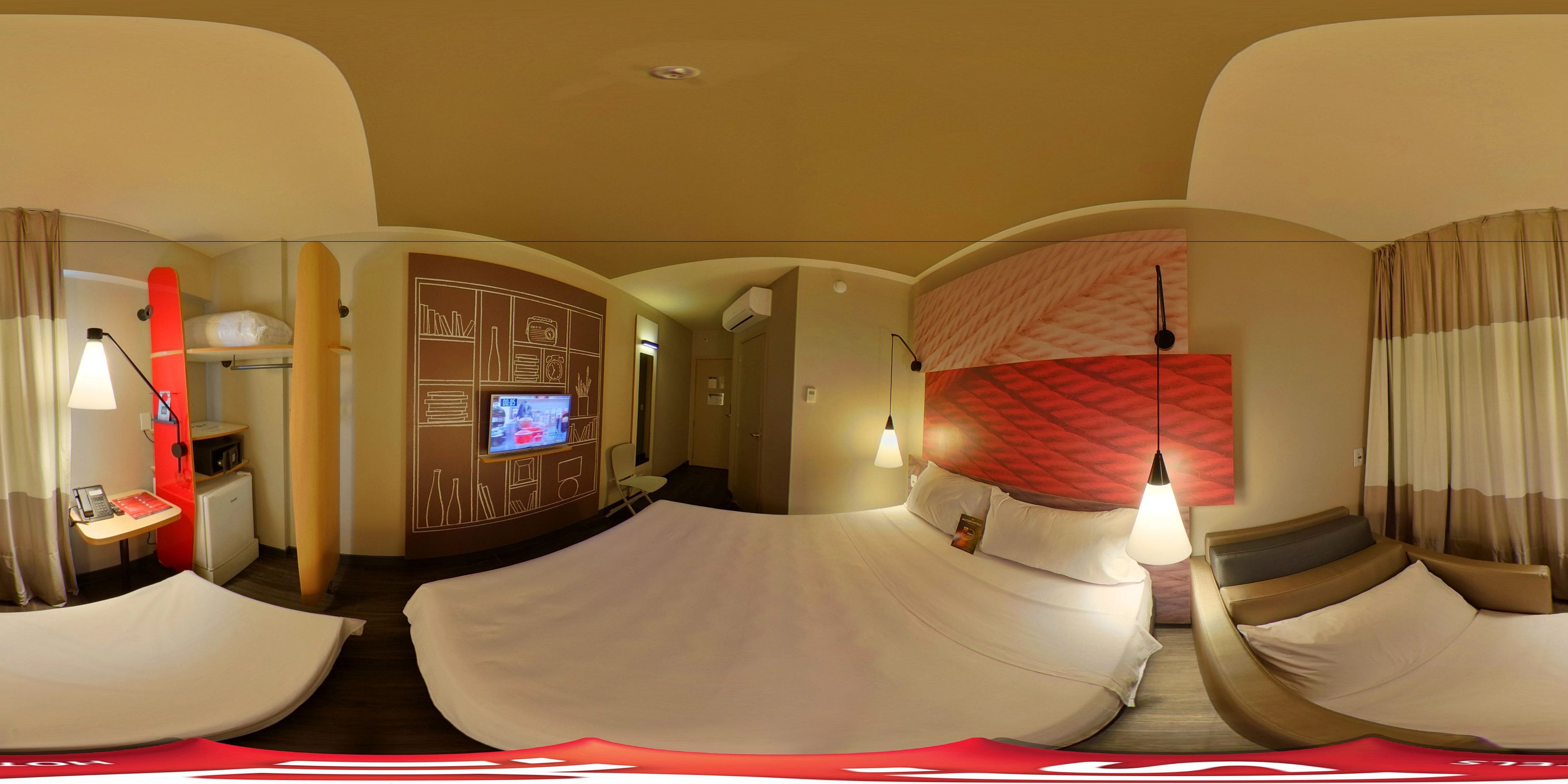 Quarto de hotel com cama, escrivaninha com abajur, televisão e parede com um padrão vermelho.