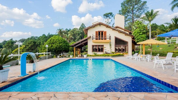 Uma piscina com um elemento aquático azul, rodeada por um alpendre com espreguiçadeiras brancas e uma casa ao fundo.