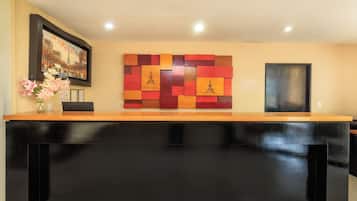 Uma receção com balcão preto, um quadro emoldurado com vista urbana, uma obra de arte abstrata colorida e um vaso com flores.