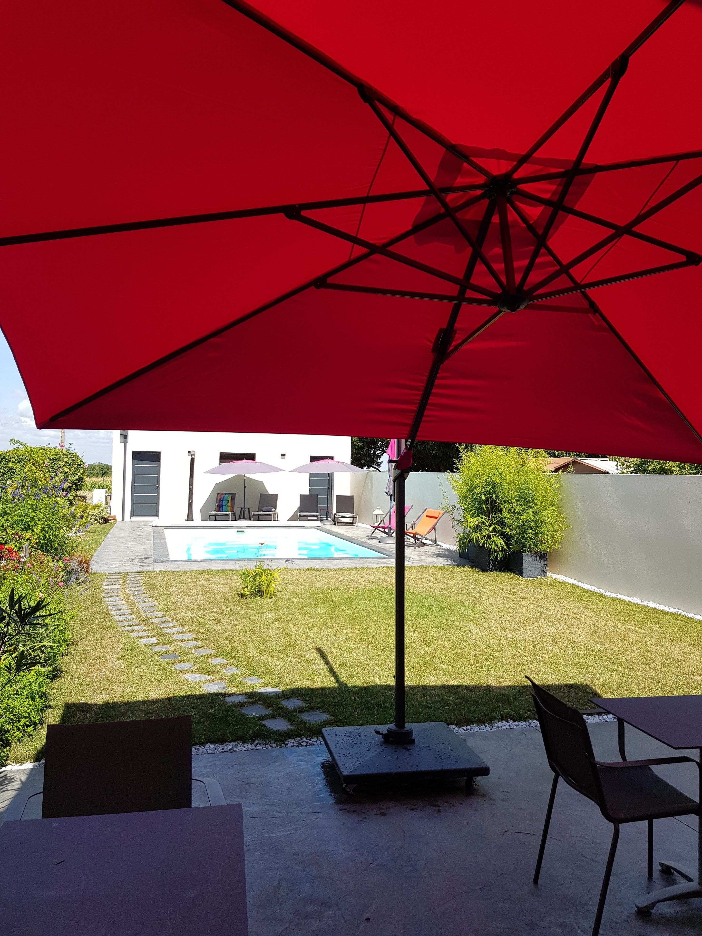 Uma área de pátio com um guarda-sol vermelho, uma piscina e um edifício branco ao fundo.