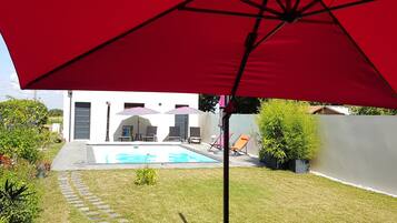 Uma área de pátio com um guarda-sol vermelho, uma piscina e um edifício branco ao fundo.