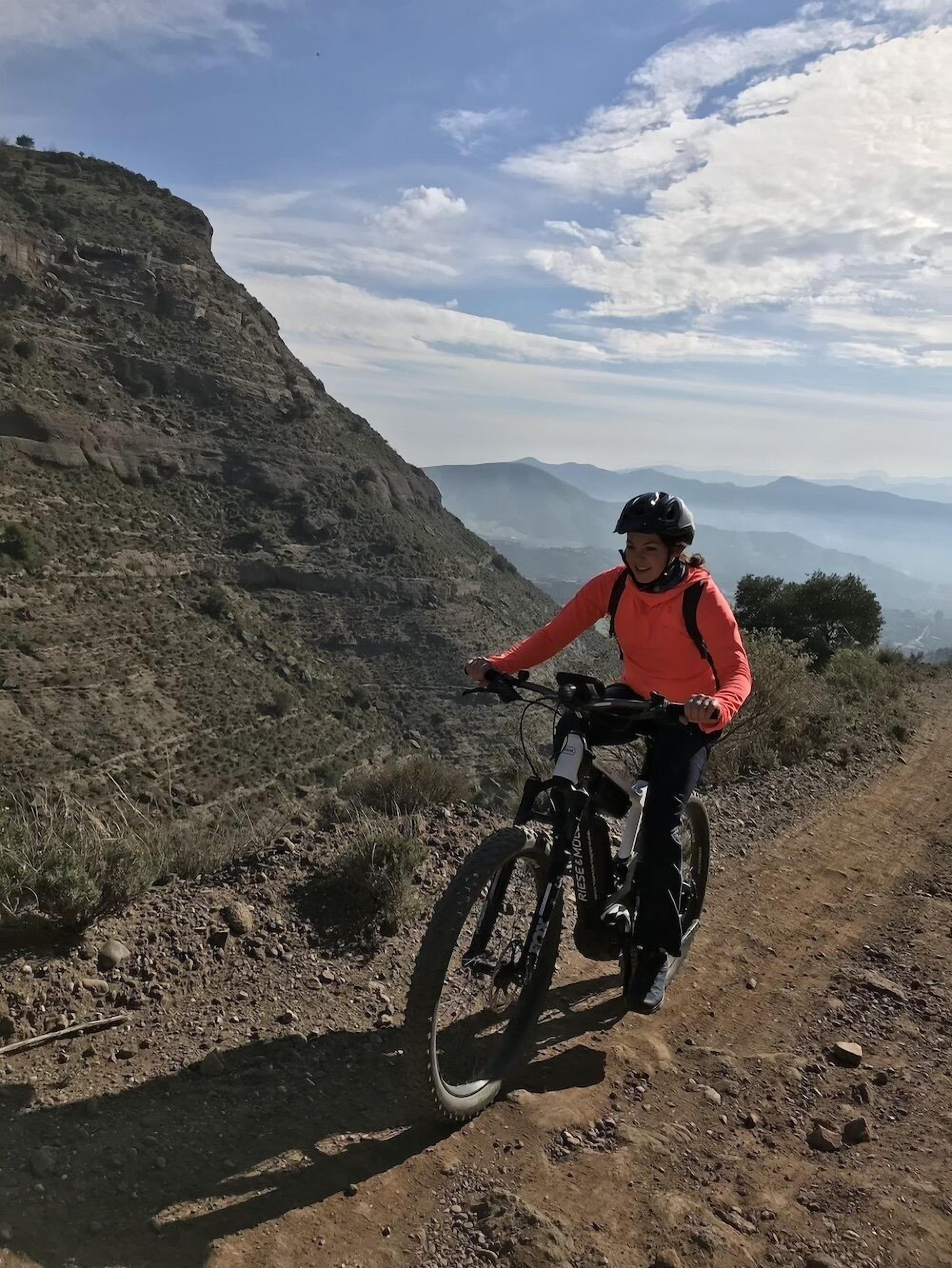 Uma pessoa andando de bicicleta de montanha em uma trilha de terra, com uma paisagem montanhosa ao fundo.