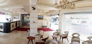 Um saguão de hotel com balcão de recepção, área de estar, lustres e uma grande janela com texto.