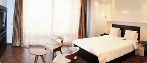 Quarto de hotel com uma cama grande, duas cadeiras, uma mesinha e uma mesa de cabeceira com abajur.